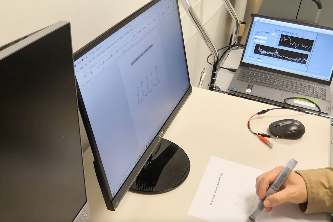 Photo | Hand holding a pen in front of two computer monitors | UCLA