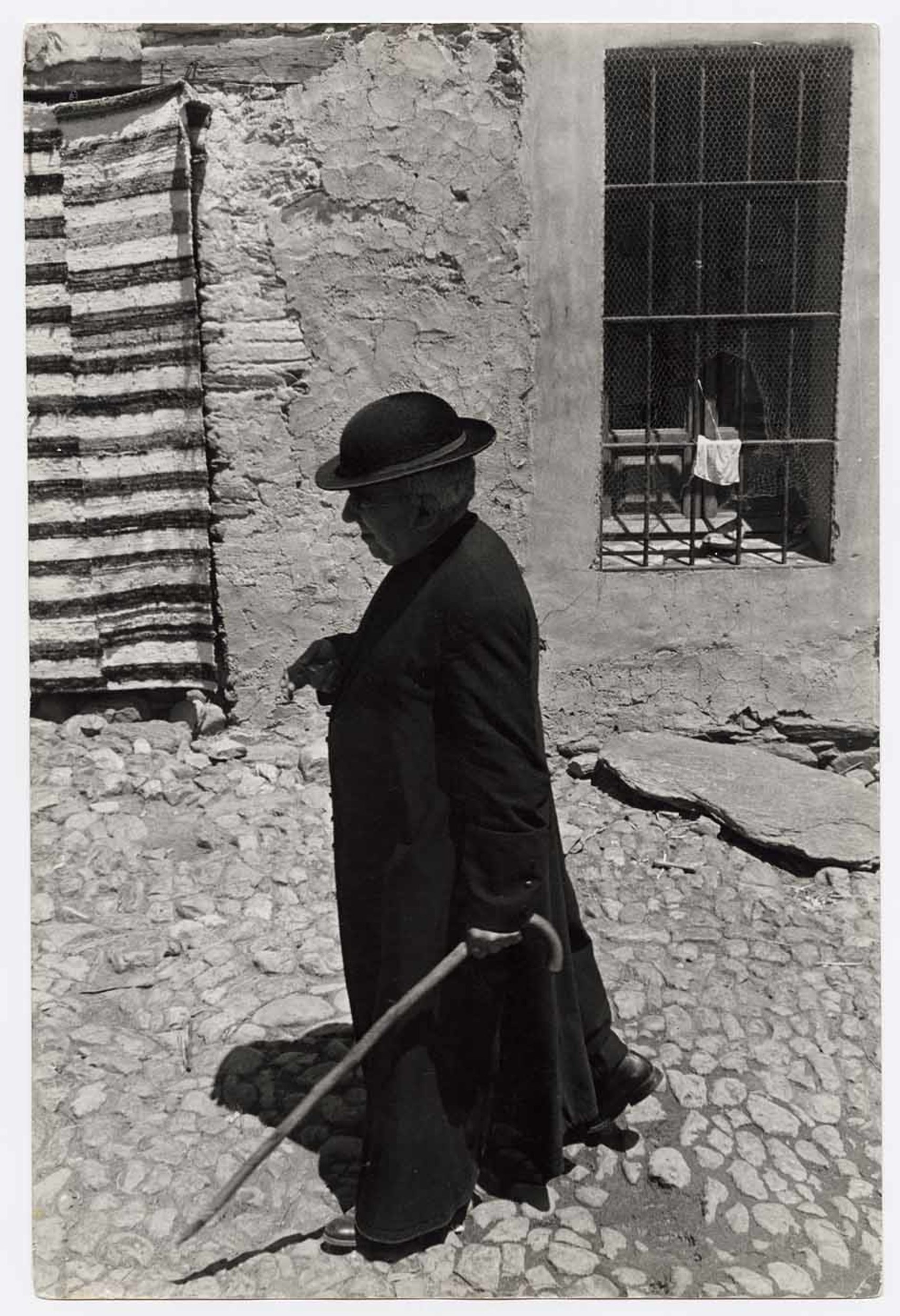 "Senor Cura" [priest, Don Manuel, walking with cane past barred window ...
