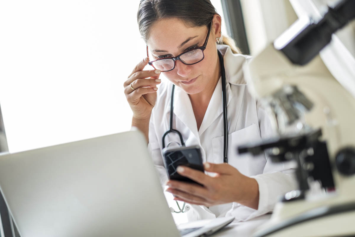 Female doctor adjusting eyeglasses while using mobile phone in ...
